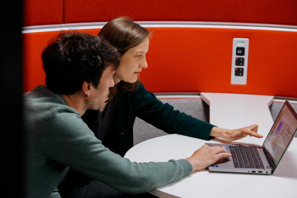 A woman with shoulder length light brown hair and a man with dark brown hair sitting at a table together looking at a laptop.