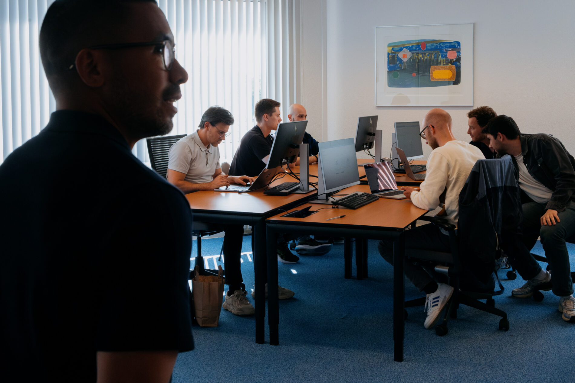 At left, a man with round glasses stands in the foreground; in the background, six men sitting at a large desk with computers are working together.