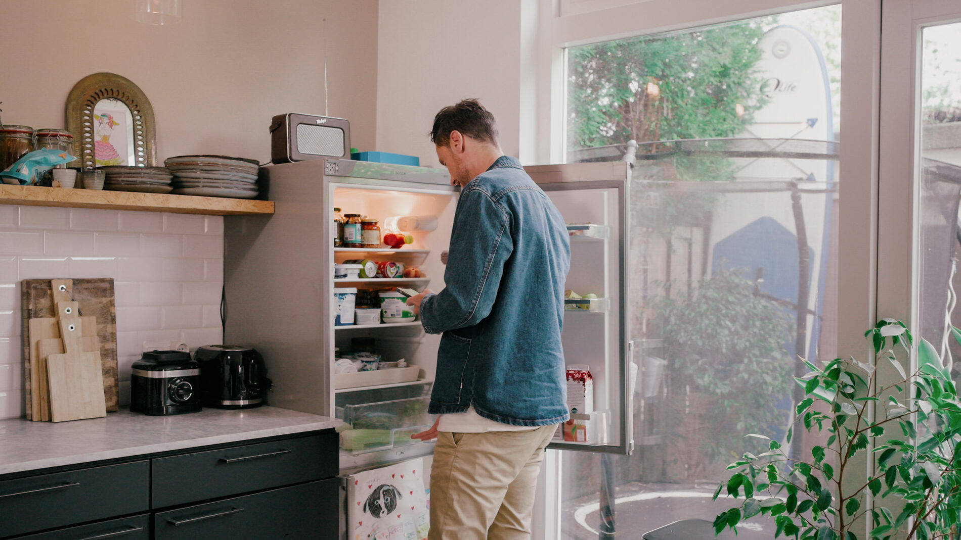 A brown-haired man in a blue shirt standing in front of an open refrigerator with a smartphone in his hand.