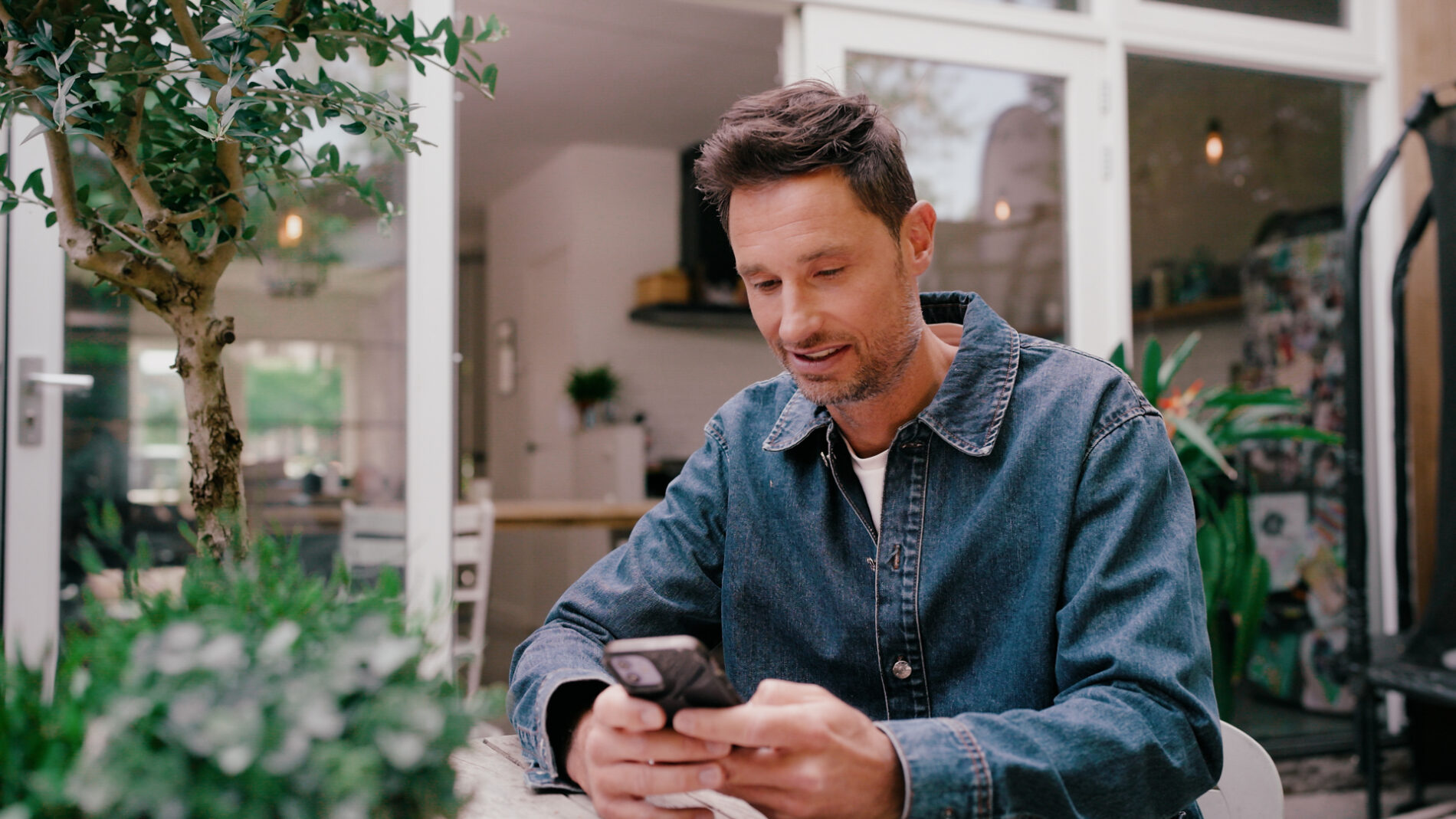 A brown-haired man in a blue shirt sitting at a table looking at his smartphone.