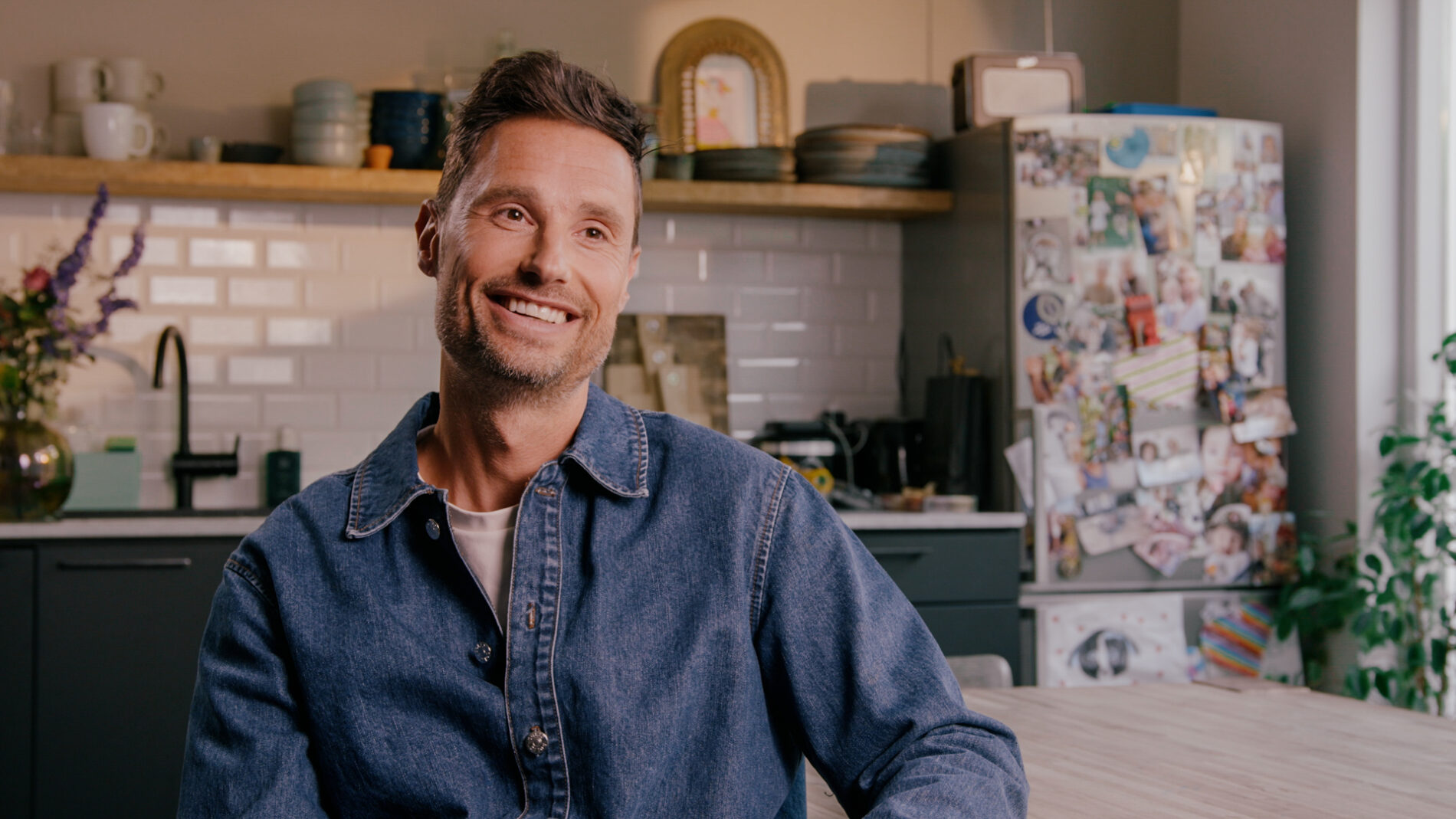 A smiling brown-haired man seated at a table with a refrigerator, kitchen counter and sink in the background.